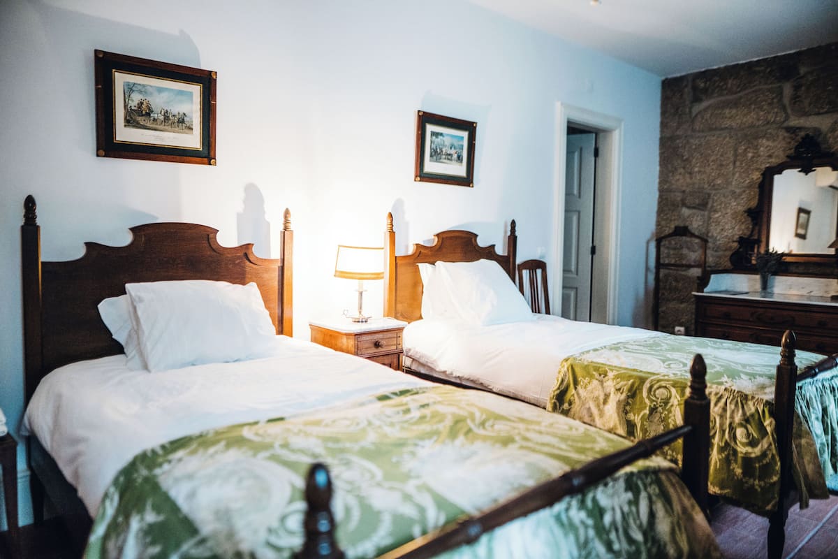 Quarto com duas camas de madeira, mesa de cabeceira com abajur, quadros na parede e parede de pedra.