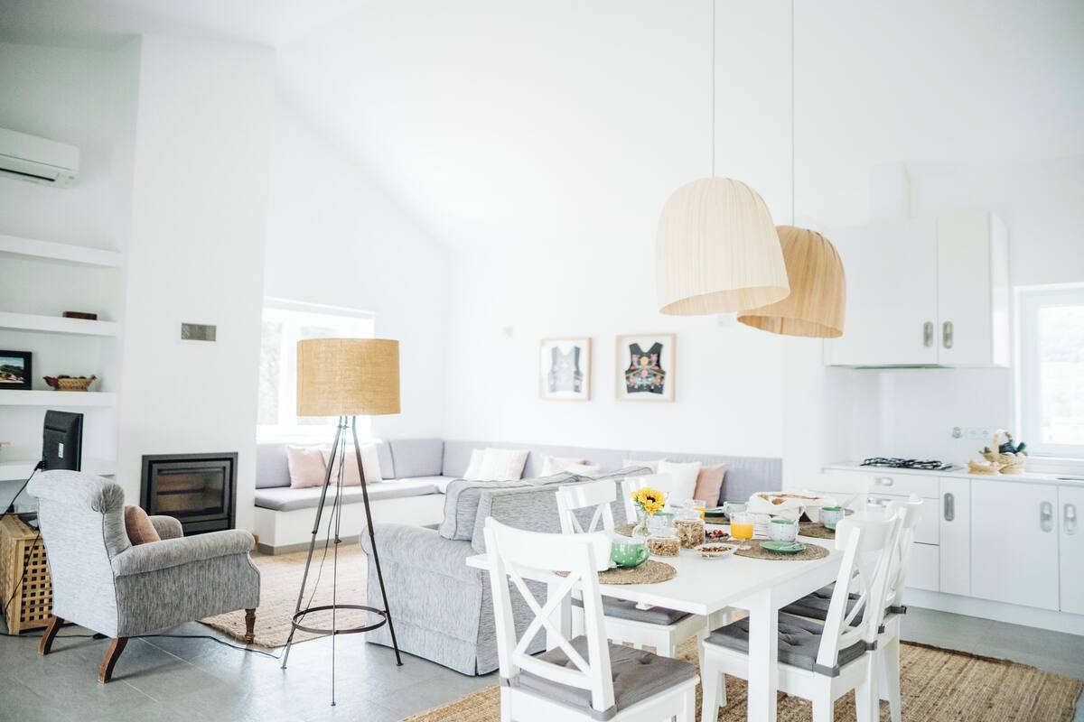 Uma sala de estar clara e moderna com sofá, mesa de jantar e cadeiras. Há um abajur, lareira e obras de arte nas paredes.