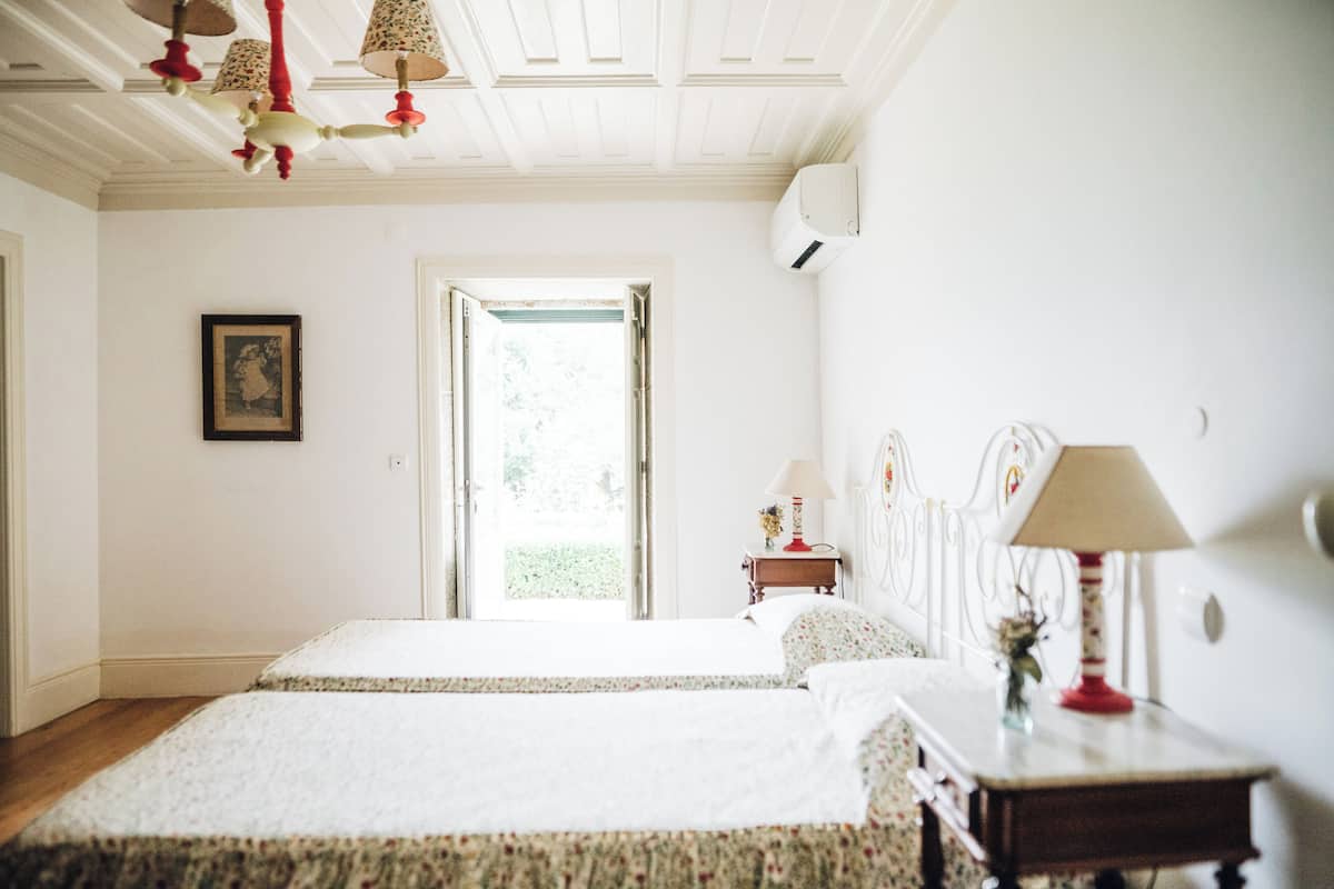 Quarto com uma cama grande, mesa de cabeceira com abajur, quadro na parede e janela com vista para área verde.