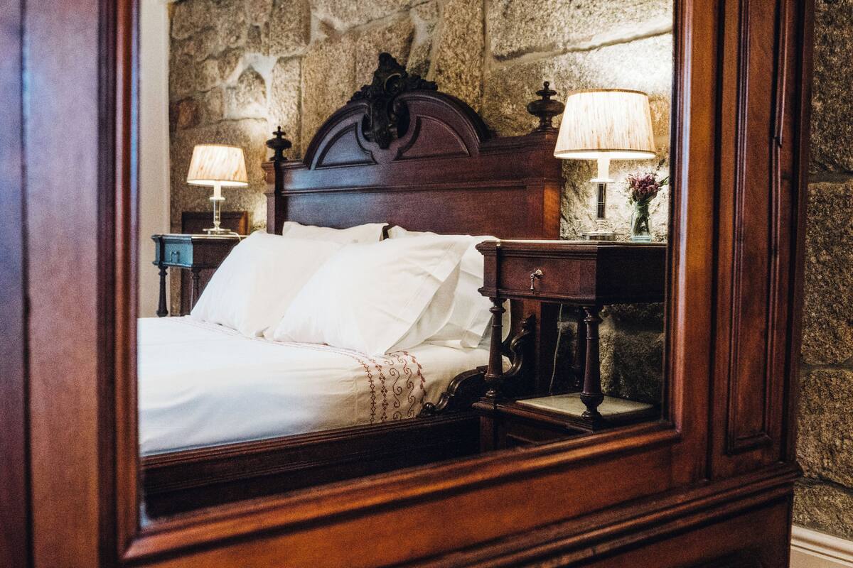 Uma cama de madeira com lençóis brancos, duas mesinhas de cabeceira com luminárias e uma parede de pedra ao fundo.