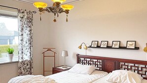 Quarto de hotel com cama, mesa de cabeceira, luminária, janela com cortinas e vista para o verde lá fora.
