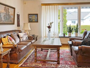 Sala de estar com sofá de couro marrom, mesa de centro de madeira e tapete estampado. Há uma janela com cortinas, um abajur e um vaso com flores.