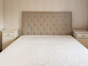 Uma cama com cabeceira estofada, duas mesas de cabeceira e uma parede lisa.