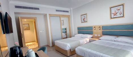 Quarto de hotel com duas camas, uma escrivaninha, uma cadeira, uma televisão e uma porta para um corredor.