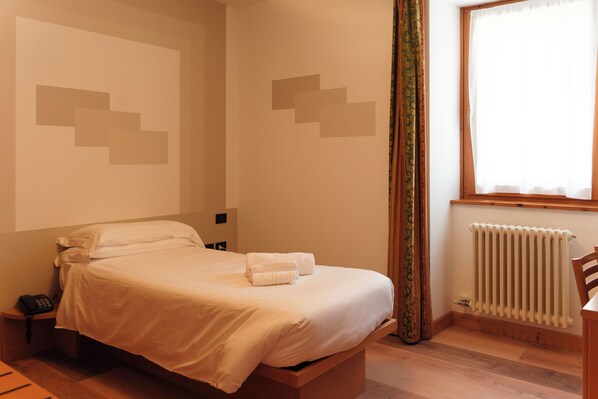 Cama bem arrumada com lençóis brancos e duas toalhas dobradas, telefone ao lado da cama, mesa de cabeceira de madeira, janela com moldura de madeira e radiador na parede.