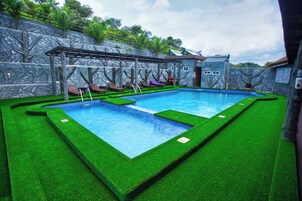 Piscina com duas áreas distintas, rodeada por relva artificial e paredes de pedra.