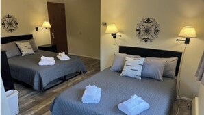 Quarto de hotel com duas camas, cada uma com colcha cinza e toalhas brancas. Há luminárias fixadas na parede, uma de cada lado das camas.