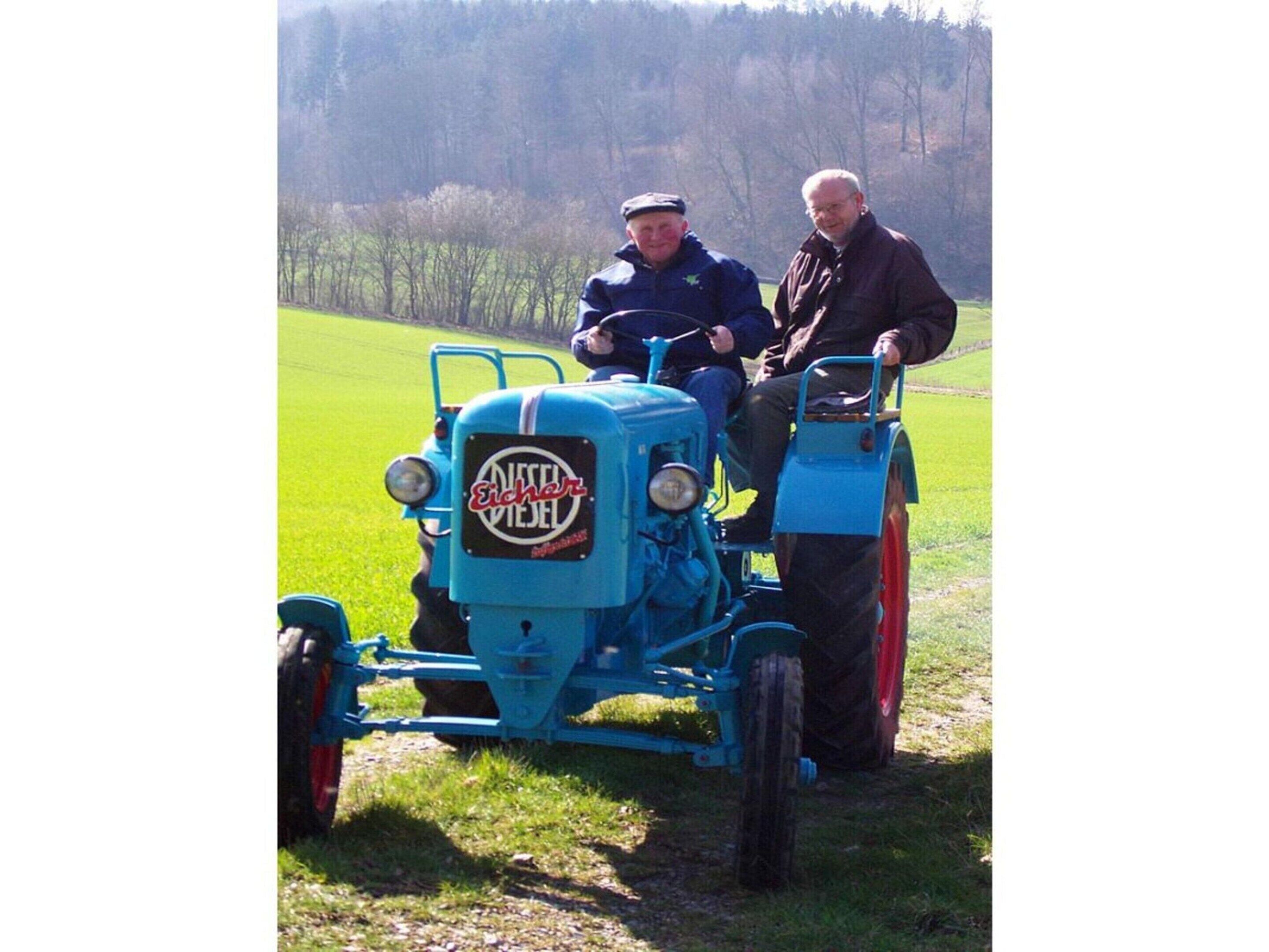 Duas pessoas em um trator vintage azul com a inscrição "Eicher", estacionado em um campo gramado com árvores ao fundo.