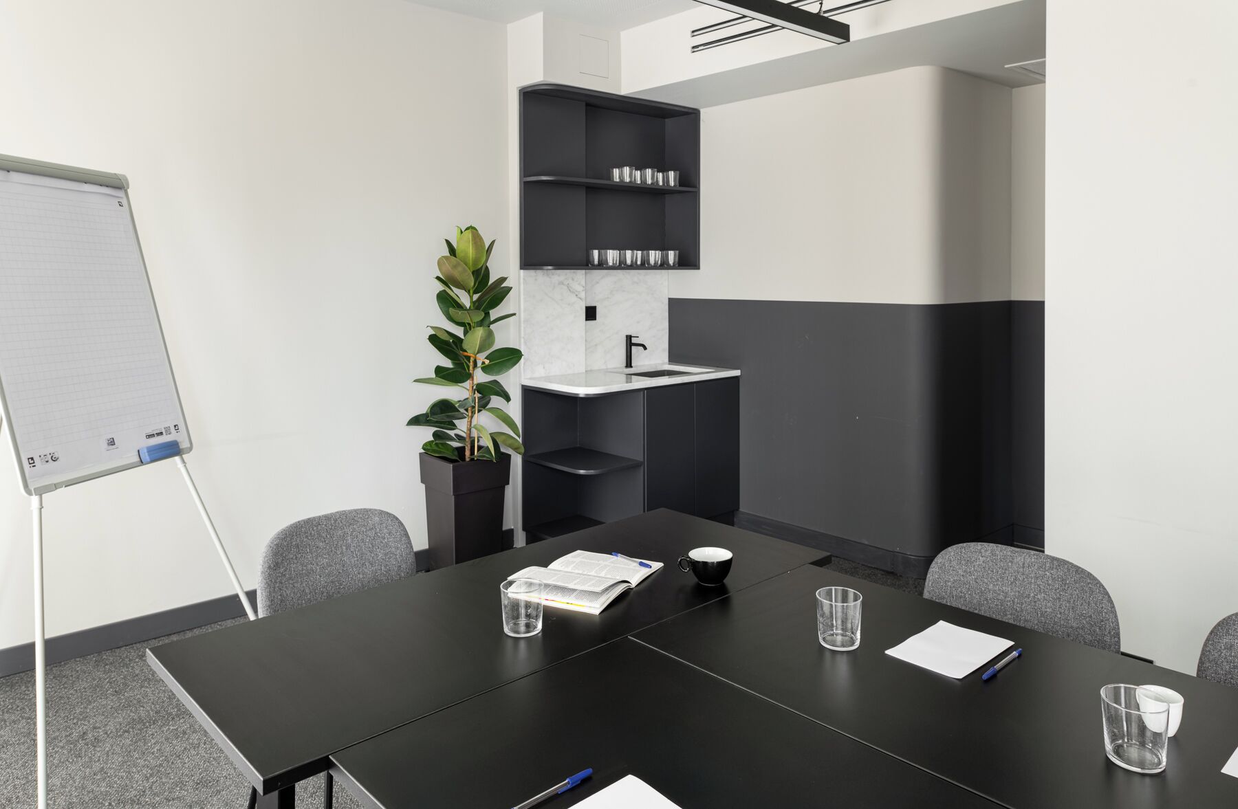 Uma sala de conferências com uma mesa preta, cadeiras, um quadro branco, uma planta, uma estante com copos e uma pia.