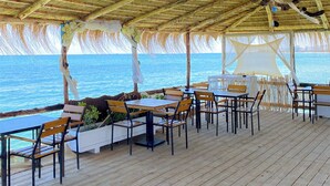 Uma área de refeições ao ar livre com mesas e cadeiras de madeira, um telhado de palha e vista para o mar.