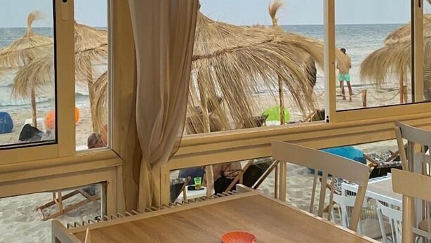 Um restaurante à beira-mar com mesa e cadeiras de madeira, grandes janelas com cortinas e guarda-sóis de palha visíveis pelas janelas.