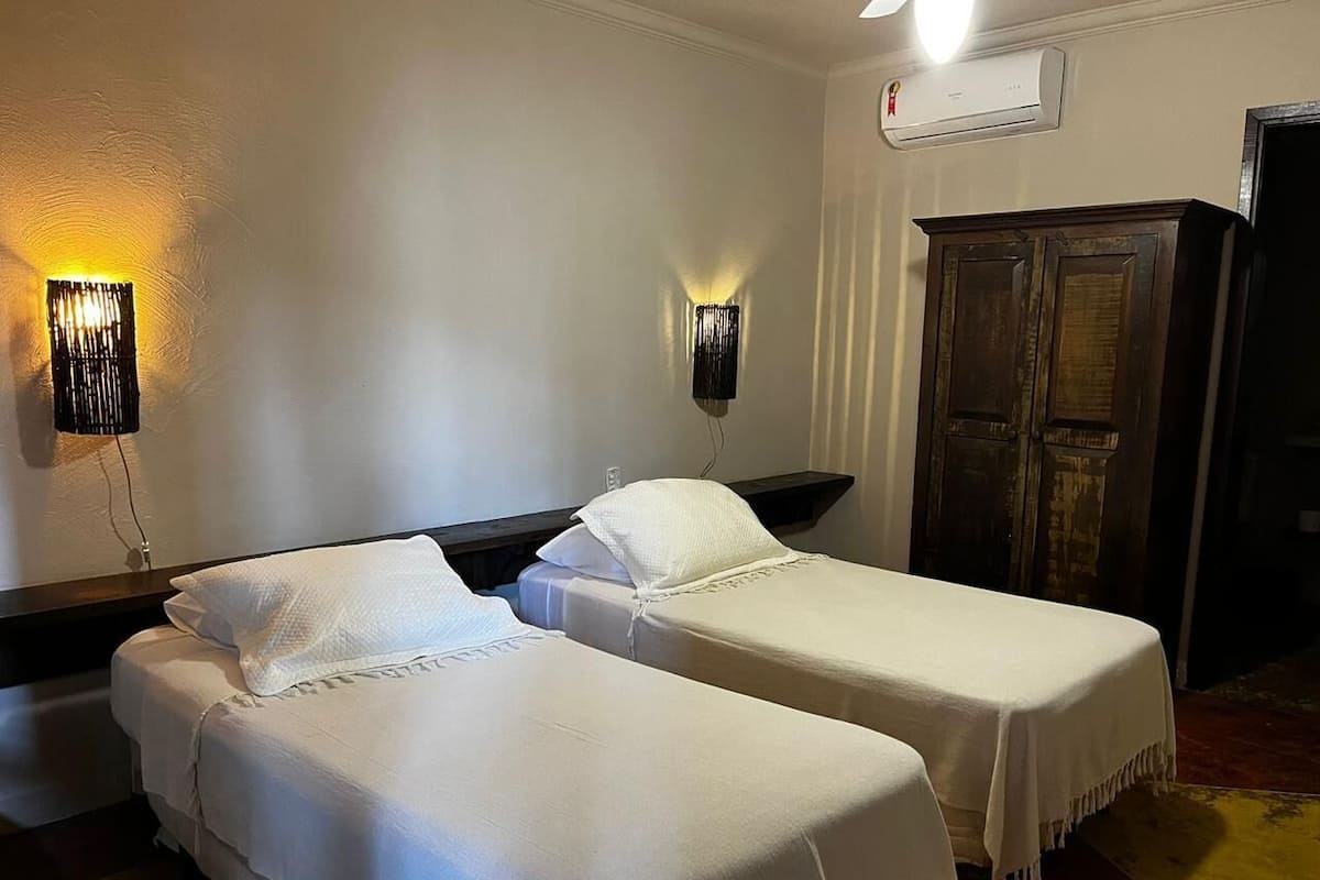 Quarto de hotel com duas camas, uma mesa de cabeceira, uma luminária na parede e um guarda-roupa.
