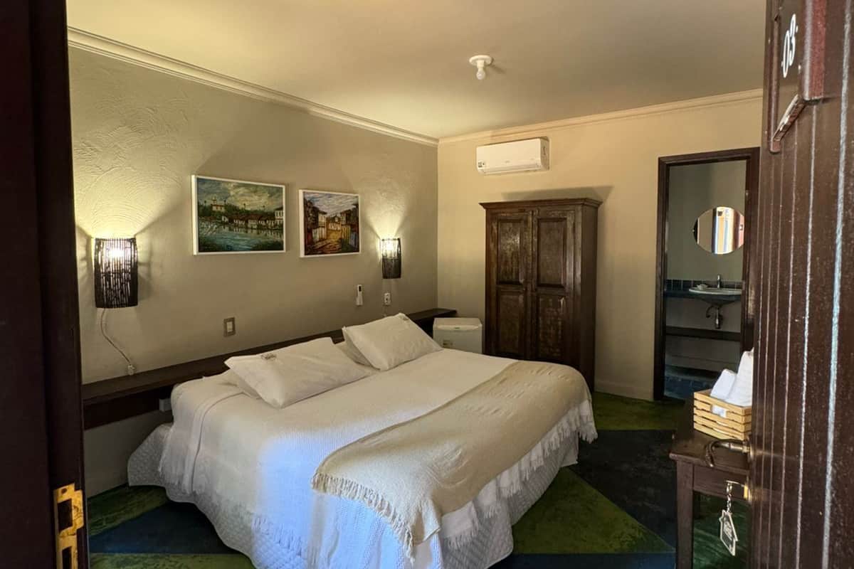 Quarto de hotel com uma cama grande, duas luminárias fixadas na parede, um guarda-roupa de madeira e uma pequena mesa com um espelho.