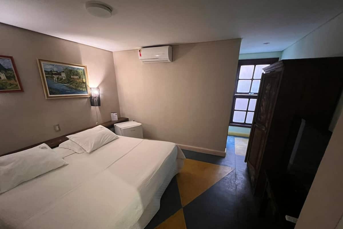 Quarto de hotel com uma cama, uma mesa de cabeceira, ar condicionado fixado na parede e uma janela.