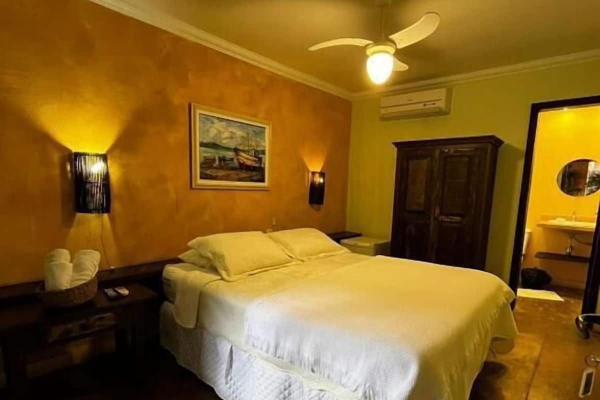 Quarto de hotel com cama, mesa de cabeceira, luminária, pintura na parede e guarda-roupa de madeira.