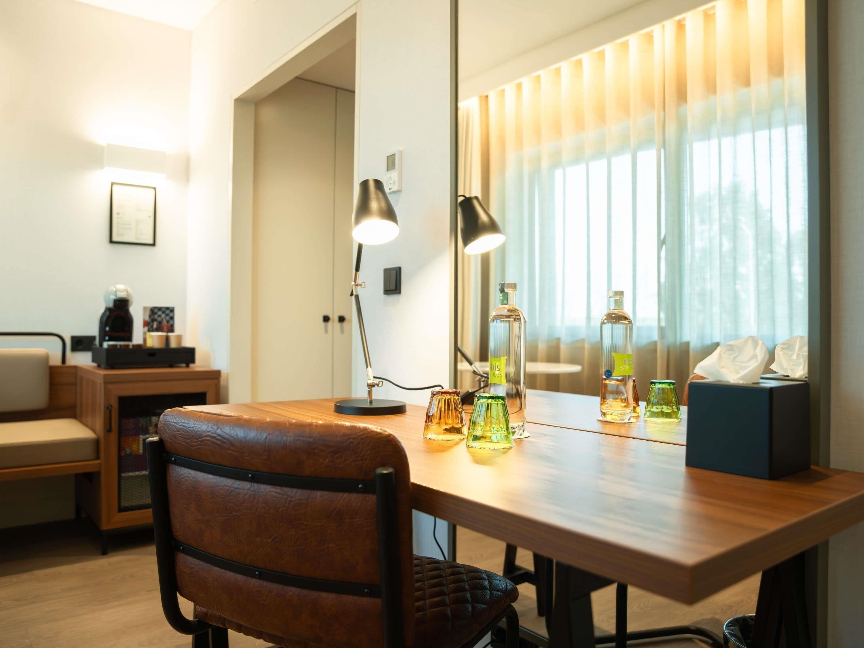 Quarto de hotel com escrivaninha de madeira, uma poltrona de couro, mesa de cabeceira com abajur e janela com persianas.