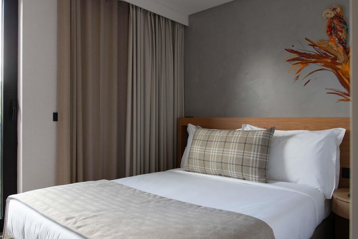 Quarto de hotel com cama, cortinas, cabeceira e um quadro na parede.