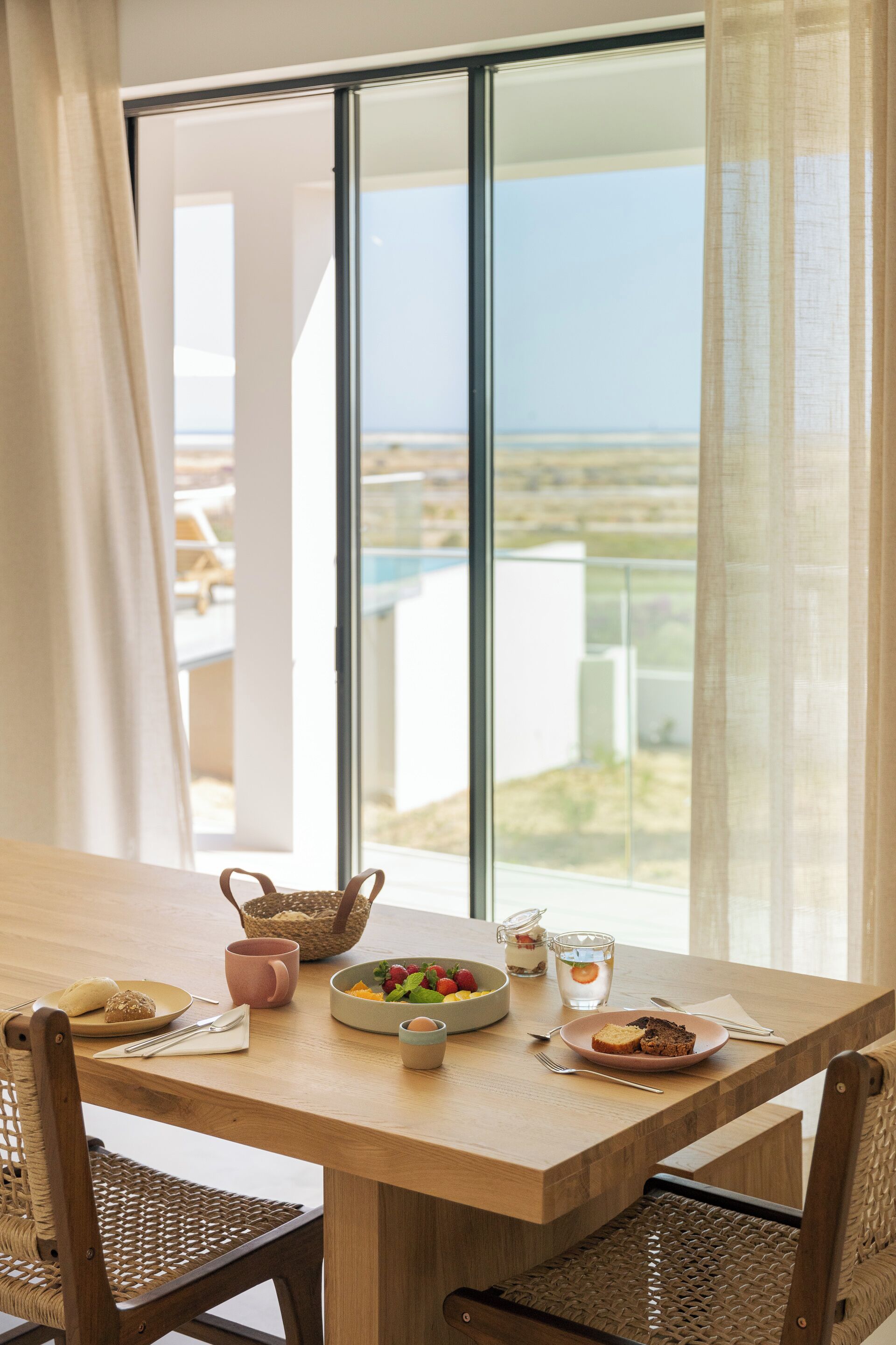 Mesa de jantar com vista para o mar através de uma porta de vidro, cadeiras de vime e uma tigela de frutas.