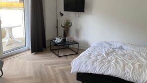 Quarto de hotel com cama, televisão fixada na parede, pequena mesa com abajur e planta, e vista para o exterior através de uma porta de correr.