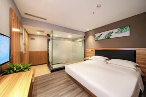 Quarto de hotel com cama, escrivaninha, televisão, área de chuveiro e um quadro na parede.