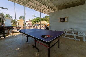 Uma mesa de pingue-pongue com raquete e bolas, um alvo de dardos e área de estar externa com guarda-sóis.