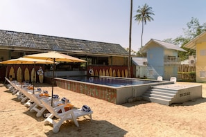 Uma área à beira da piscina com espreguiçadeiras e guarda-sóis, piscina e um edifício com telhado inclinado.