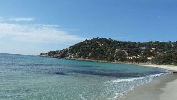 Uma praia com águas turquesa cristalinas, areal arenoso e um morro com edifícios.