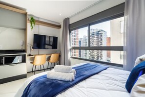 Quarto de hotel moderno com uma cama grande, uma escrivaninha com TV de tela plana, duas cadeiras, micro-ondas e vista para a cidade através de amplas janelas.