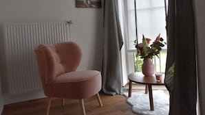 Quarto com uma cadeira rosa, uma mesa pequena com um vaso de flores e uma janela com cortinas.