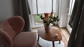 Um quarto com uma cadeira rosa, uma pequena mesa de madeira com um vaso de flores e uma janela com cortinas.