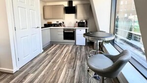 Cozinha compacta com eletrodomésticos modernos, área de refeições com uma mesa e cadeira pequenas, e uma grande janela com vista para a cidade.