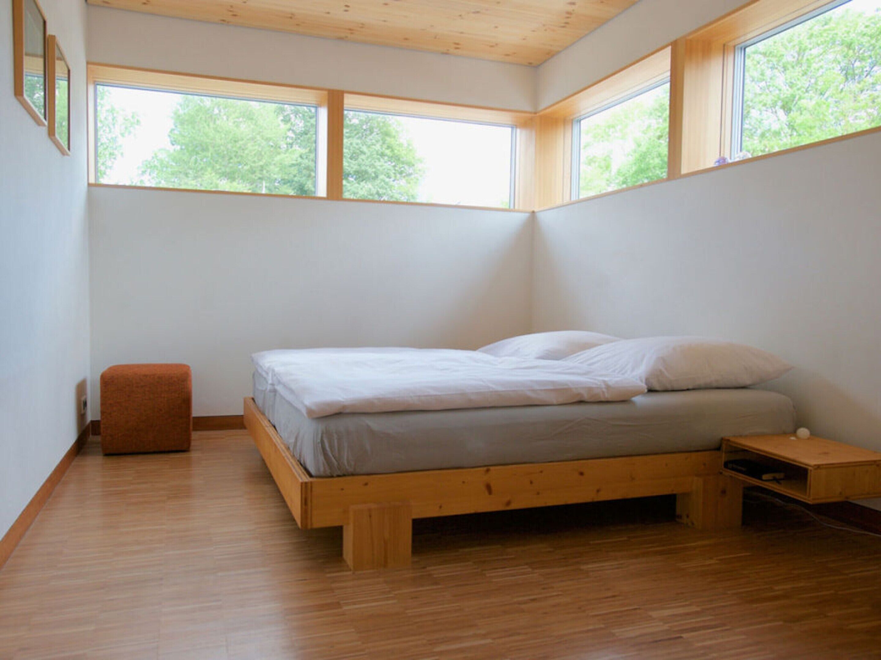 Quarto com cama de madeira, mesa de cabeceira e um pequeno banquinho. Janelas amplas oferecem iluminação natural.