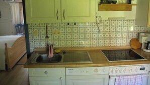 Uma cozinha com armários brancos, uma pia, um fogão e um micro-ondas. Há uma prateleira de madeira acima da pia com uma chaleira e xícaras.
