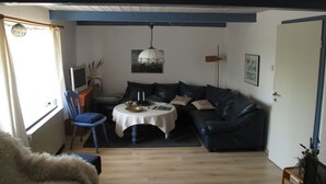 Uma sala de estar com um sofá de couro escuro, uma mesa redonda pequena com toalha branca, uma poltrona azul e um cobertor de pele no chão.