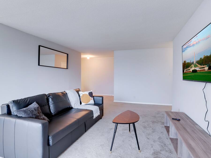 Uma sala de estar moderna com um sofá cinza, uma pequena mesa de centro de madeira e uma TV de tela plana fixada na parede.