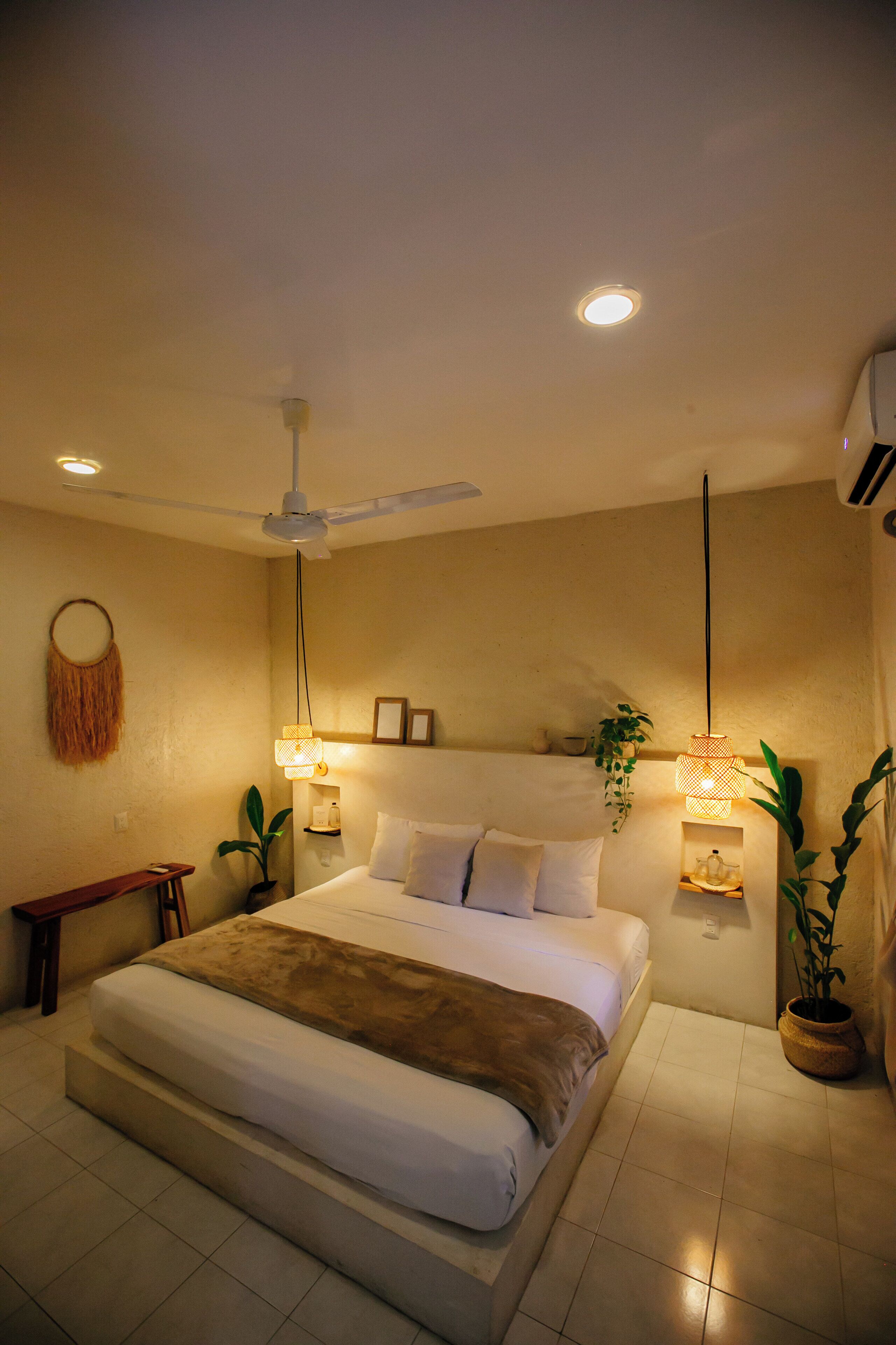 Quarto com cama, ventilador de teto, luminárias na parede e planta na mesinha de cabeceira.