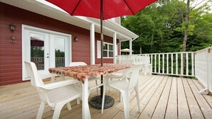 Um deck de madeira com mesa e cadeiras, um guarda-sol vermelho e um corrimão branco.