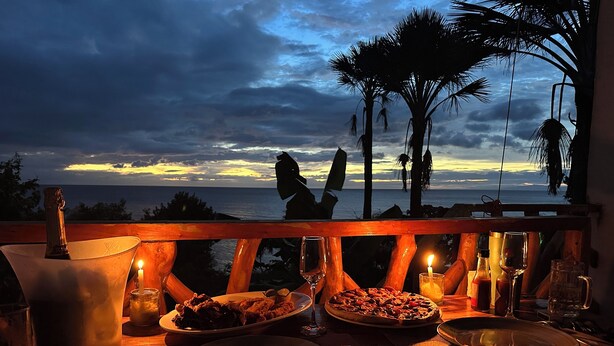 Mesa posta para jantar, com pratos de comida, copos e uma vela, com vista para um cenário oceânico ao entardecer.