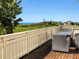 Um deck de madeira com móveis de vime, guarda-corpo branco e vista para o oceano e colinas.