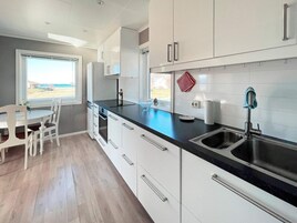 Cozinha moderna com armários brancos, bancada preta e cuba de aço inoxidável. Há uma janela com vista para a praia.