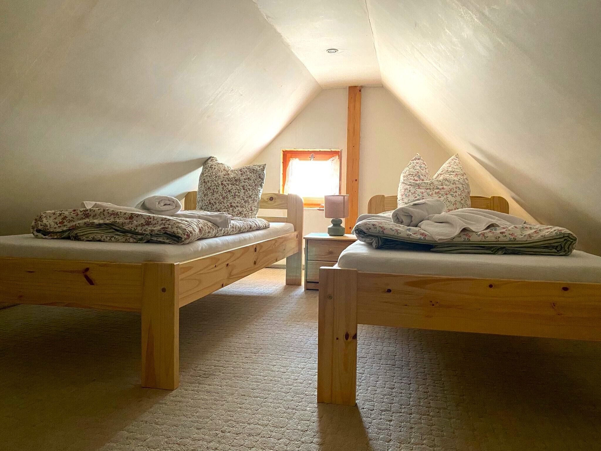 Um quarto de sótão pequeno e acolhedor, com duas camas beliches de madeira, uma mesinha e uma janela que oferece luz natural.