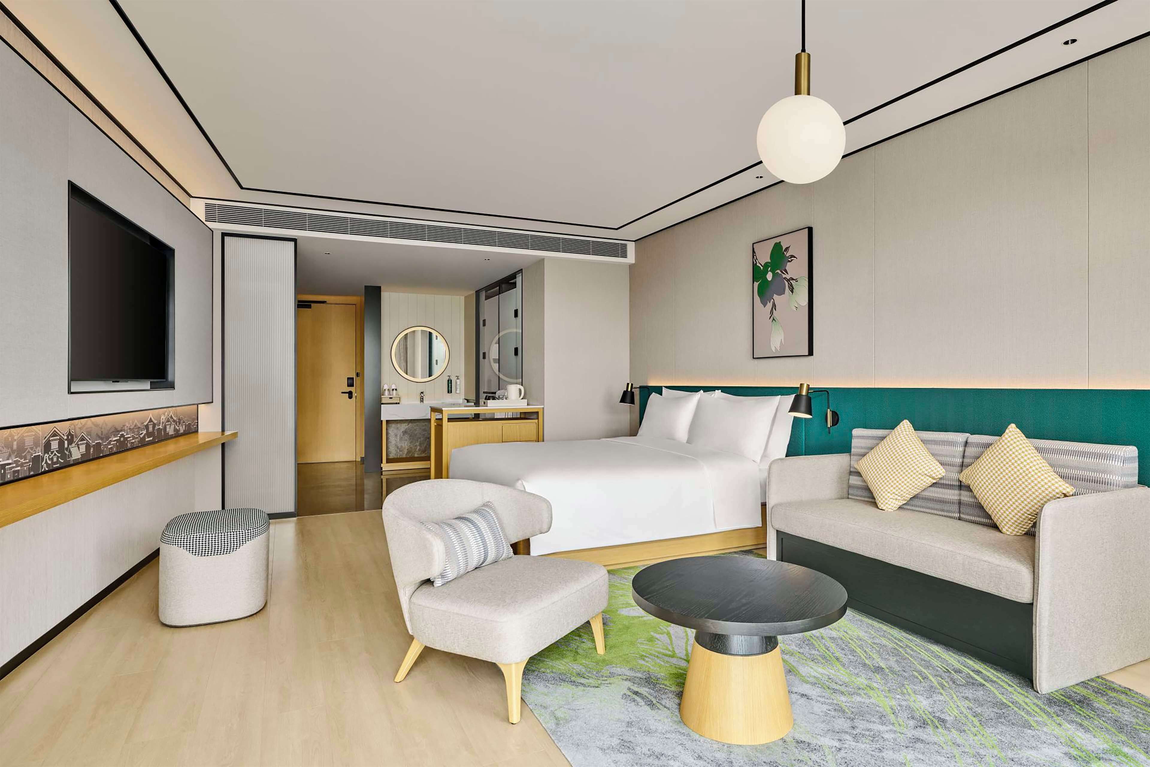 Quarto de hotel moderno com cama, sofá, poltrona e uma mesa pequena. Há uma televisão, um espelho e um quadro na parede.