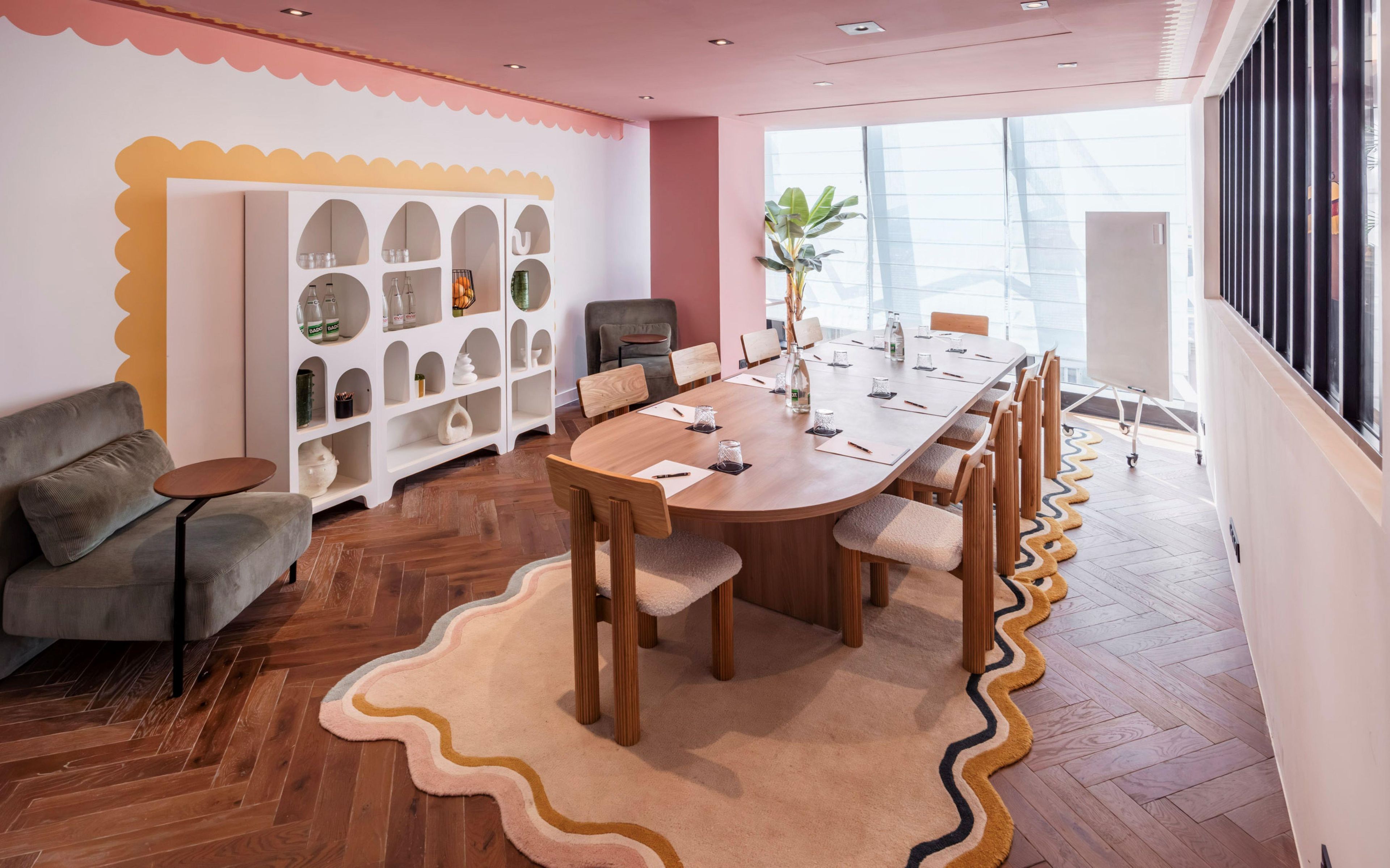 Uma sala de reunião moderna com uma mesa redonda, cadeiras de madeira, uma estante branca com objetos decorativos e uma grande janela que oferece luz natural.