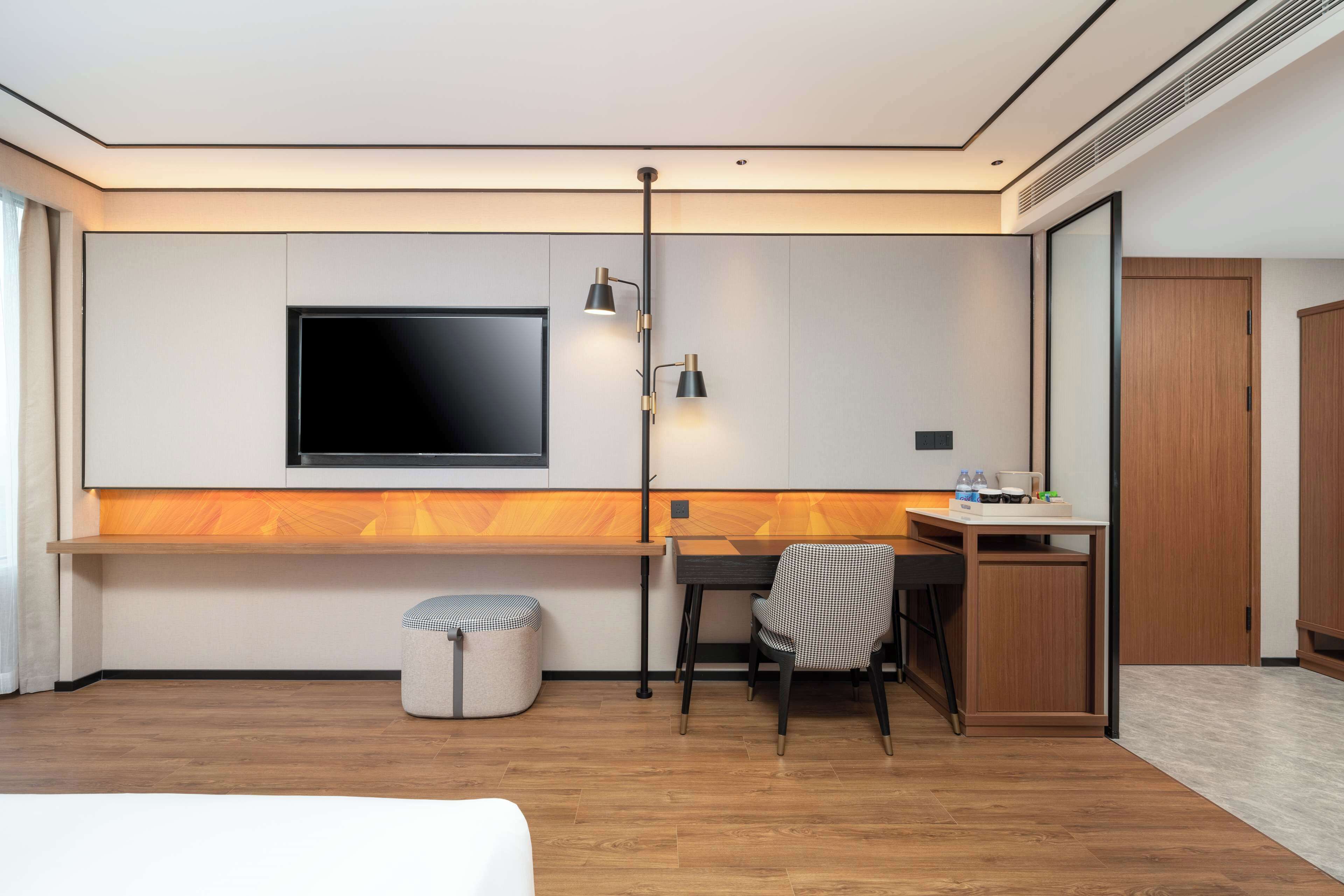 Quarto de hotel moderno com televisão de tela plana, escrivaninha com cadeira, um banquinho pequeno e um guarda-roupa.