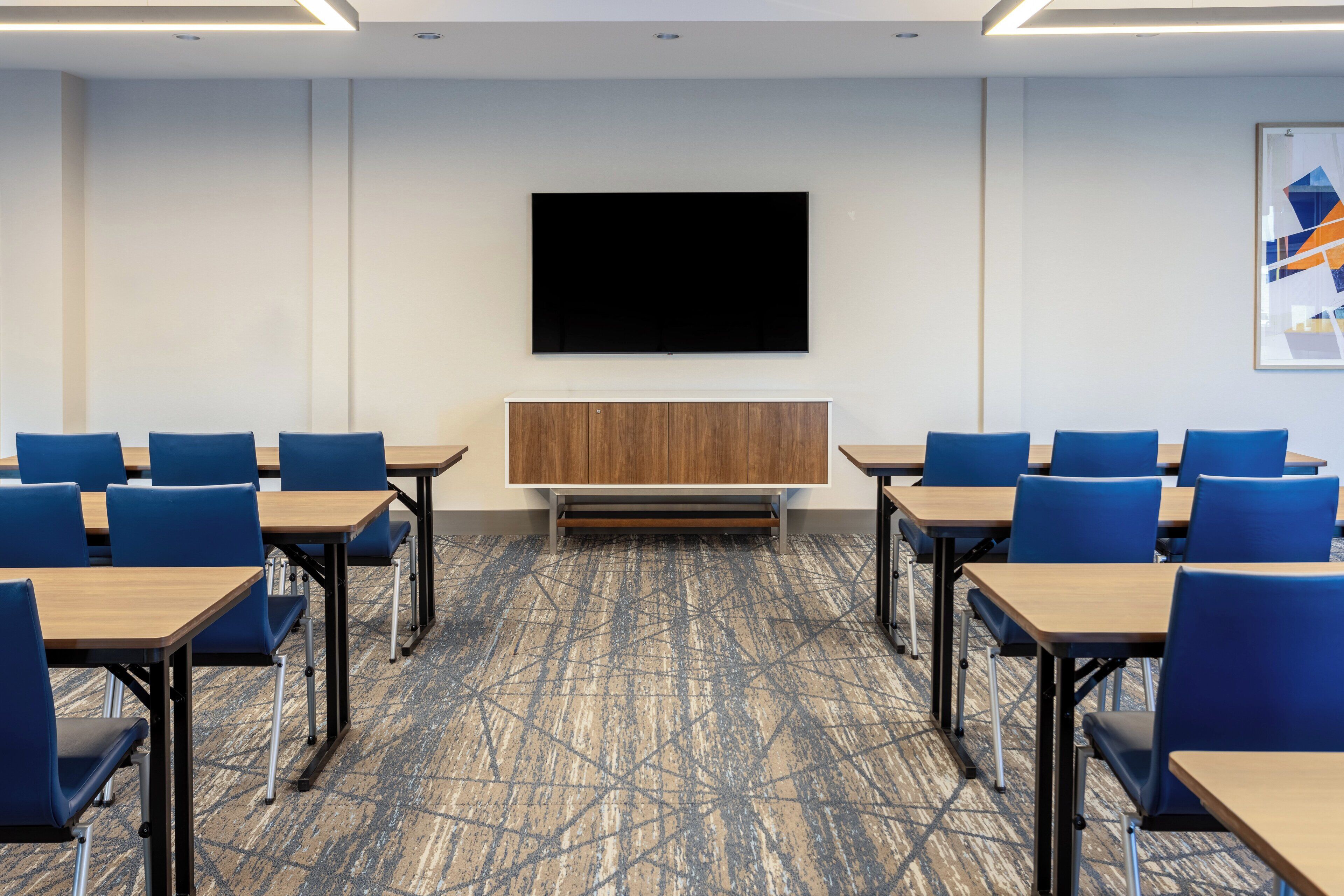 Uma sala de conferência com uma grande televisão de tela plana fixada na parede, um painel de madeira no centro, e cadeiras azuis dispostas ao redor de mesas de madeira.