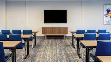 Uma sala de conferência com uma grande televisão de tela plana fixada na parede, um painel de madeira no centro, e cadeiras azuis dispostas ao redor de mesas de madeira.