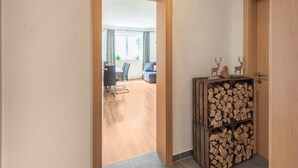 Um corredor com uma porta de madeira que dá para uma sala de jantar, com uma mesa e cadeiras. Há uma pilha de lenha encostada na parede.