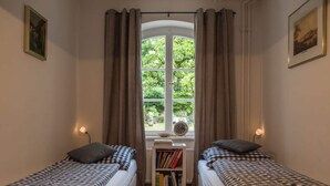 Quarto pequeno com duas camas, uma janela com cortinas, uma estante e um relógio.