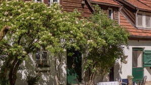 Uma casa de madeira tradicional com porta verde, um pequeno jardim com vasos de plantas e uma mesa com cadeiras do lado de fora.