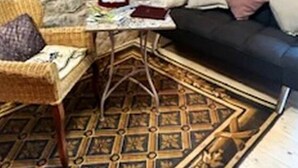 Quarto aconchegante com parede de pedra, sofá preto, cadeira de vime e tapete estampado.